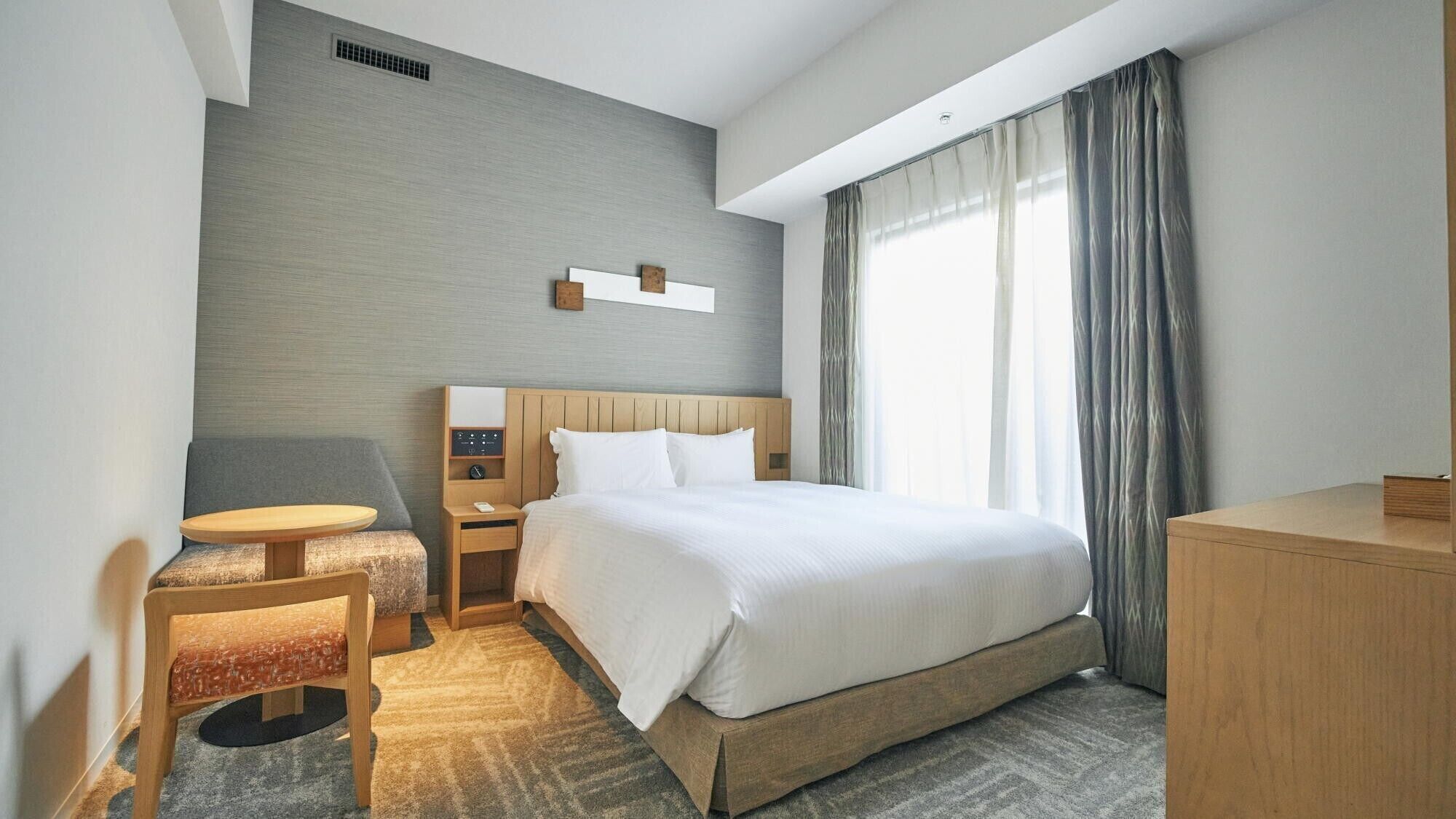 Quarto de hotel com cama, mesa de cabeceira, cadeira e escrivaninha com luminária.