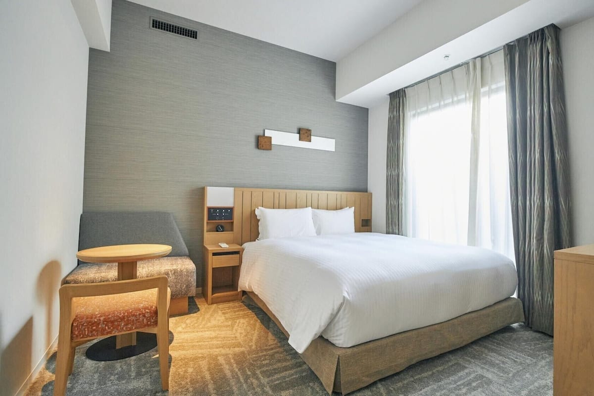 Quarto de hotel com cama, mesa de cabeceira, cadeira e escrivaninha com luminária.