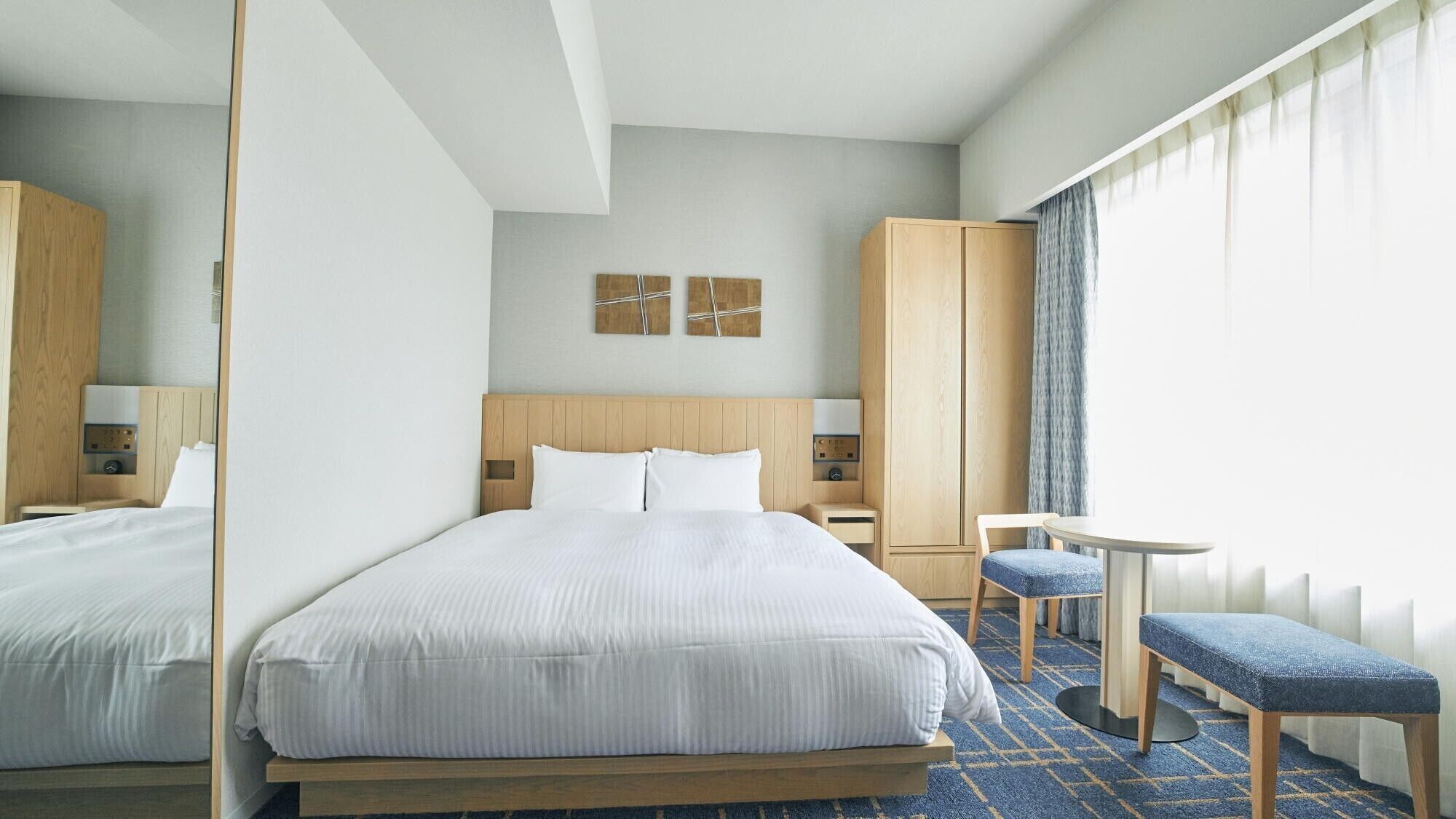 Quarto de hotel com uma cama grande, uma mesinha, uma cadeira e um guarda-roupa.