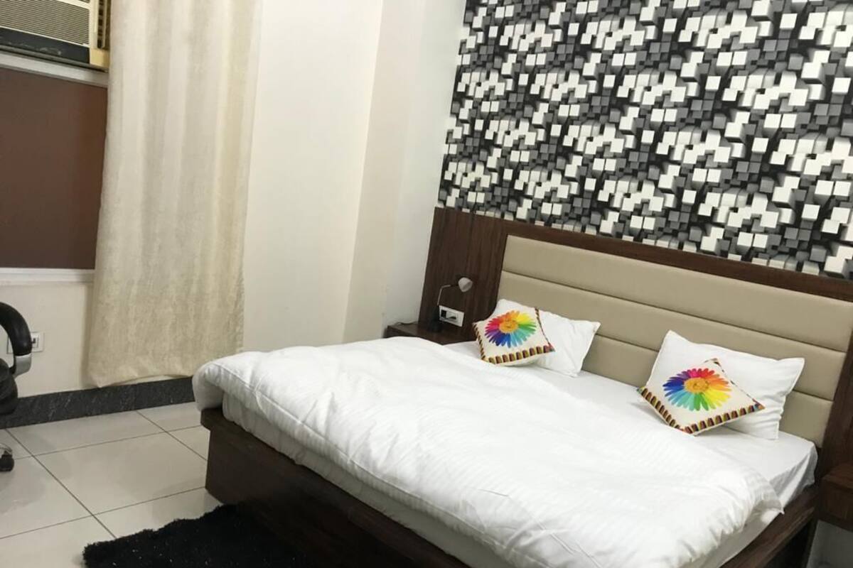 Cama bem arrumada com roupa de cama branca e duas almofadas coloridas, um tapete preto e uma parede estampada atrás da cama.