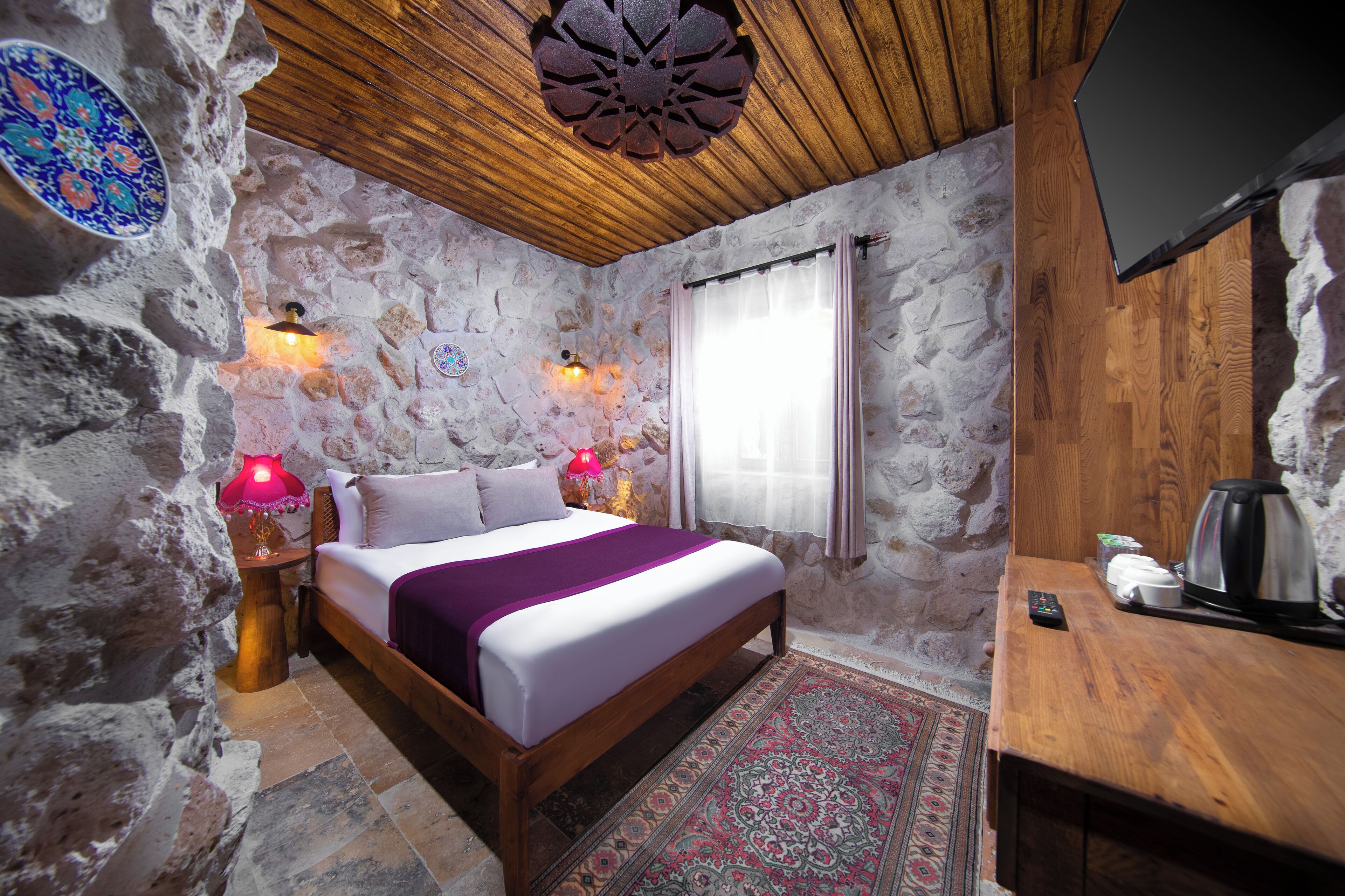 Quarto com paredes de pedra, uma cama de madeira, uma mesinha de cabeceira com abajur, uma cafeteira e uma TV sobre uma mesa de madeira.