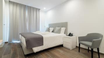 Quarto de hotel moderno com uma cama grande, uma mesinha de cabeceira com telefone e uma poltrona cinzenta.