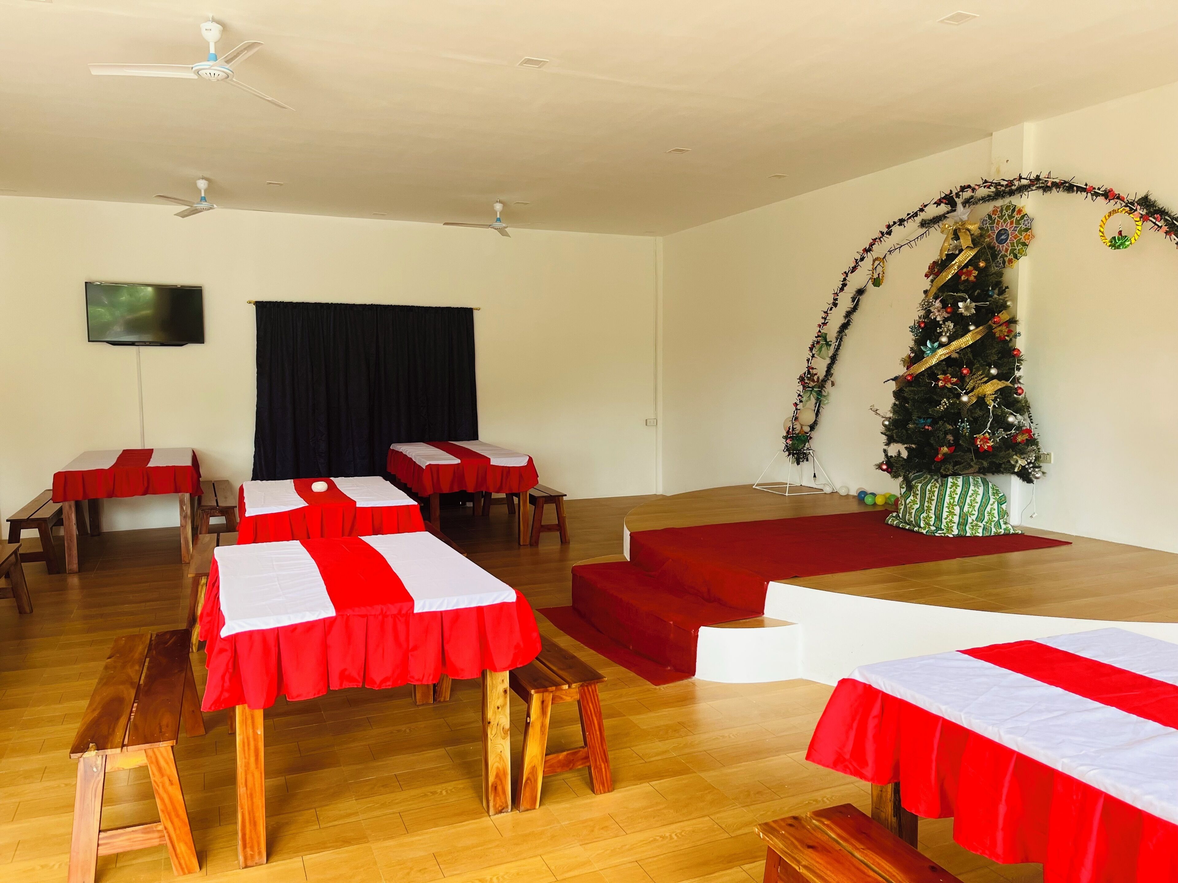 Um salão com várias mesas cobertas por toalhas vermelhas e brancas, um pinheiro de Natal e ventiladores de teto.