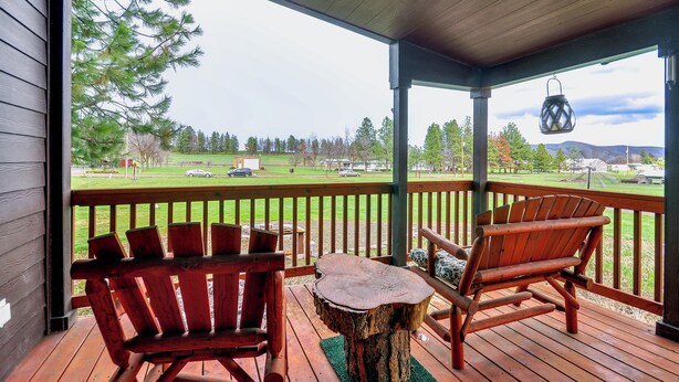 Um deck de madeira com duas cadeiras Adirondack e uma mesa pequena, com vista para uma área gramada com árvores e veículos distantes.