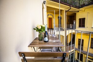 Mesa de madeira com garrafa de vinho e copos numa varanda com vista para um edifício amarelo com varandas.