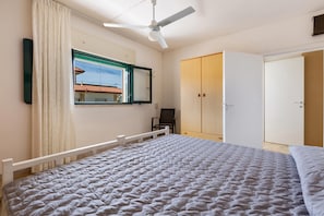 Quarto com cama, ventilador de teto, janela com vista para um edifício e guarda-roupa.