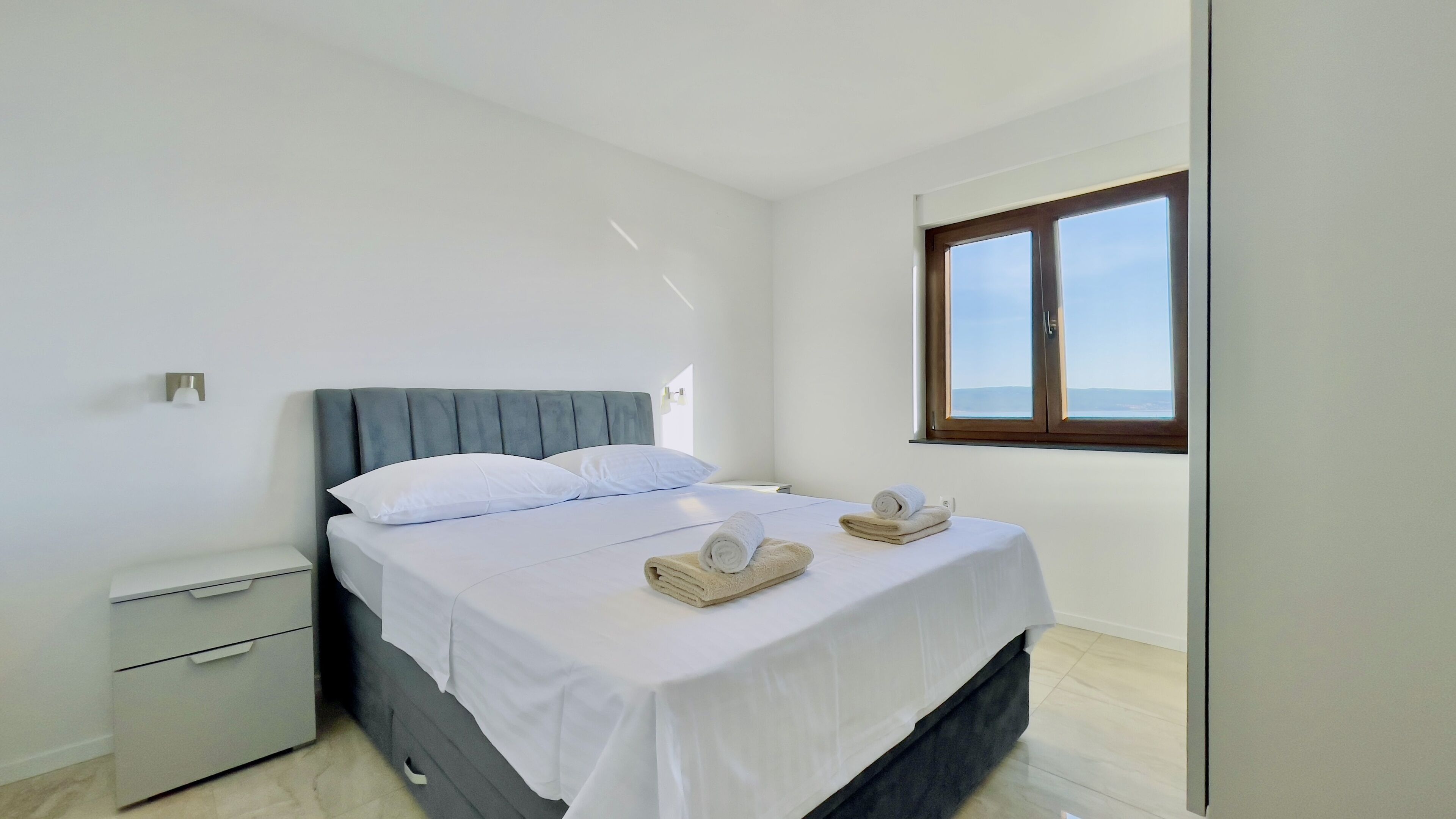 Quarto moderno com cabeceira estofada cinza, edredom branco, duas almofadas, mesa de cabeceira com abajur e janela com vista.