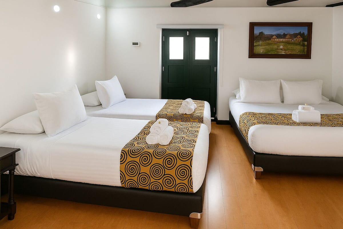 Quarto de hotel com três camas, cada uma com roupa de cama branca e colcha estampada. Há uma porta verde e um quadro na parede.