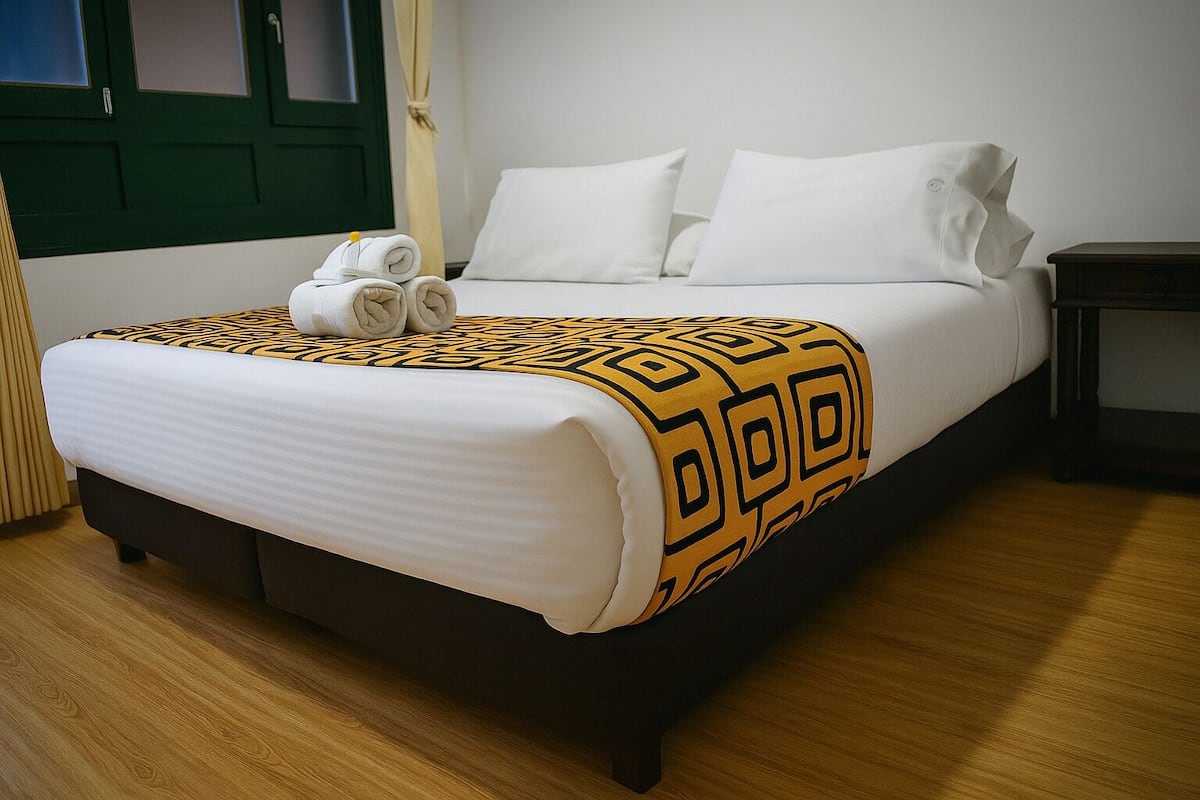 Cama bem arrumada com colcha estampada, travesseiros brancos e toalhas dobradas na cabeceira.