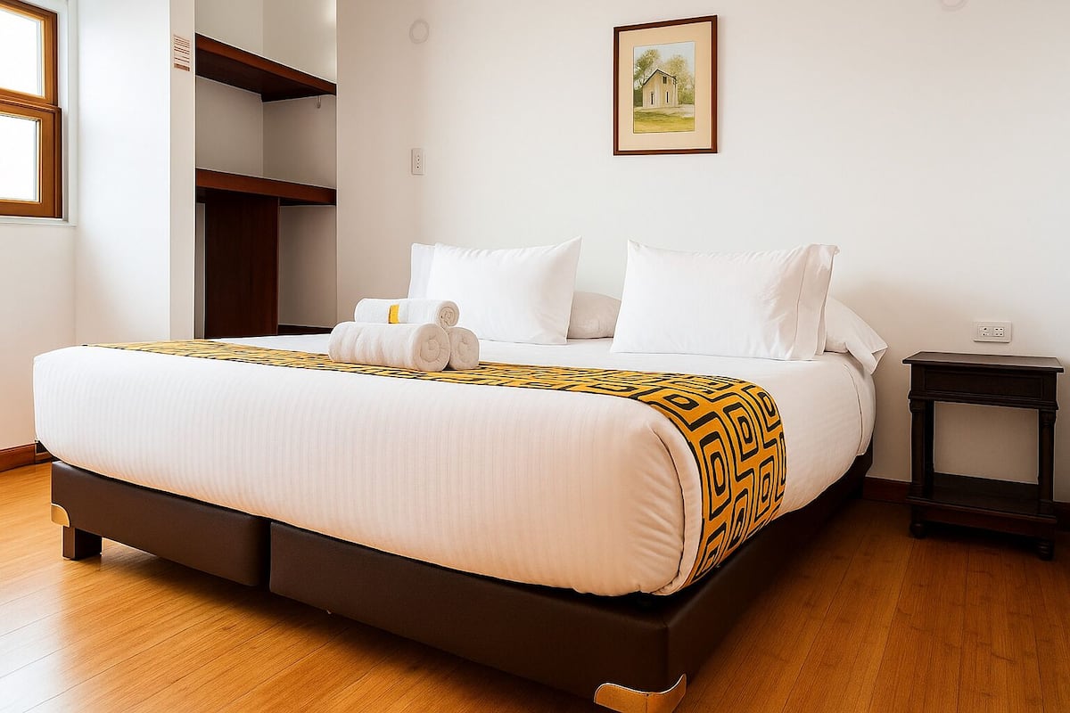 Quarto de hotel com uma cama grande, piso de madeira, mesa de cabeceira pequena e um quadro na parede.