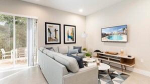 Uma sala de estar moderna com um sofá modular cinza, uma mesa de centro branca e vista para a piscina através de uma porta de vidro deslizante.