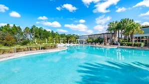 Uma piscina ampla e translúcida com área cercada, palmeiras e um edifício com varanda coberta ao fundo.