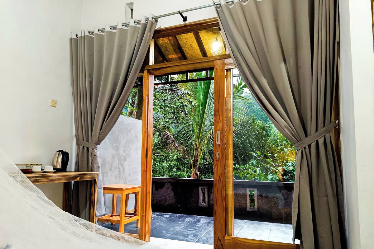 Quarto com escrivaninha de madeira, uma cadeira e um banquinho. Há uma janela com cortinas e vista para o verde lá fora.