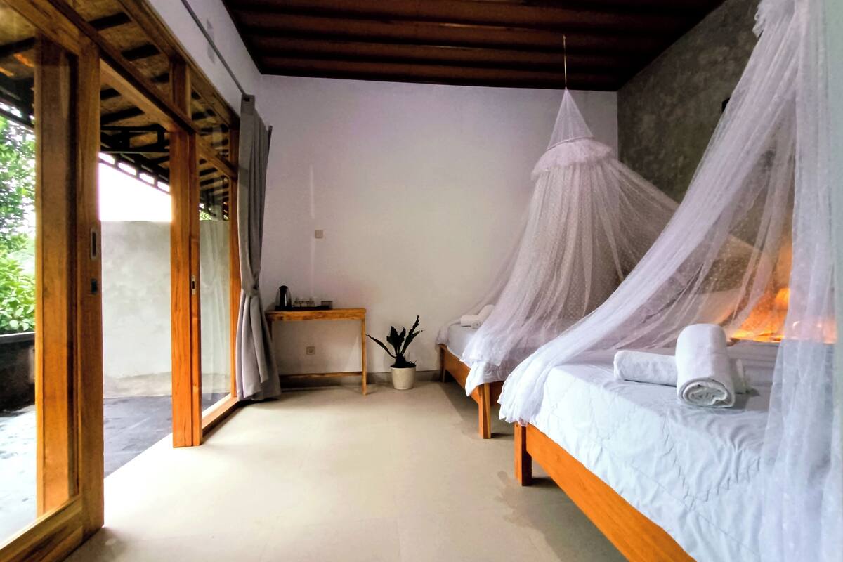 Quarto com duas camas, um banco de madeira, uma mesa pequena e um vaso com planta.