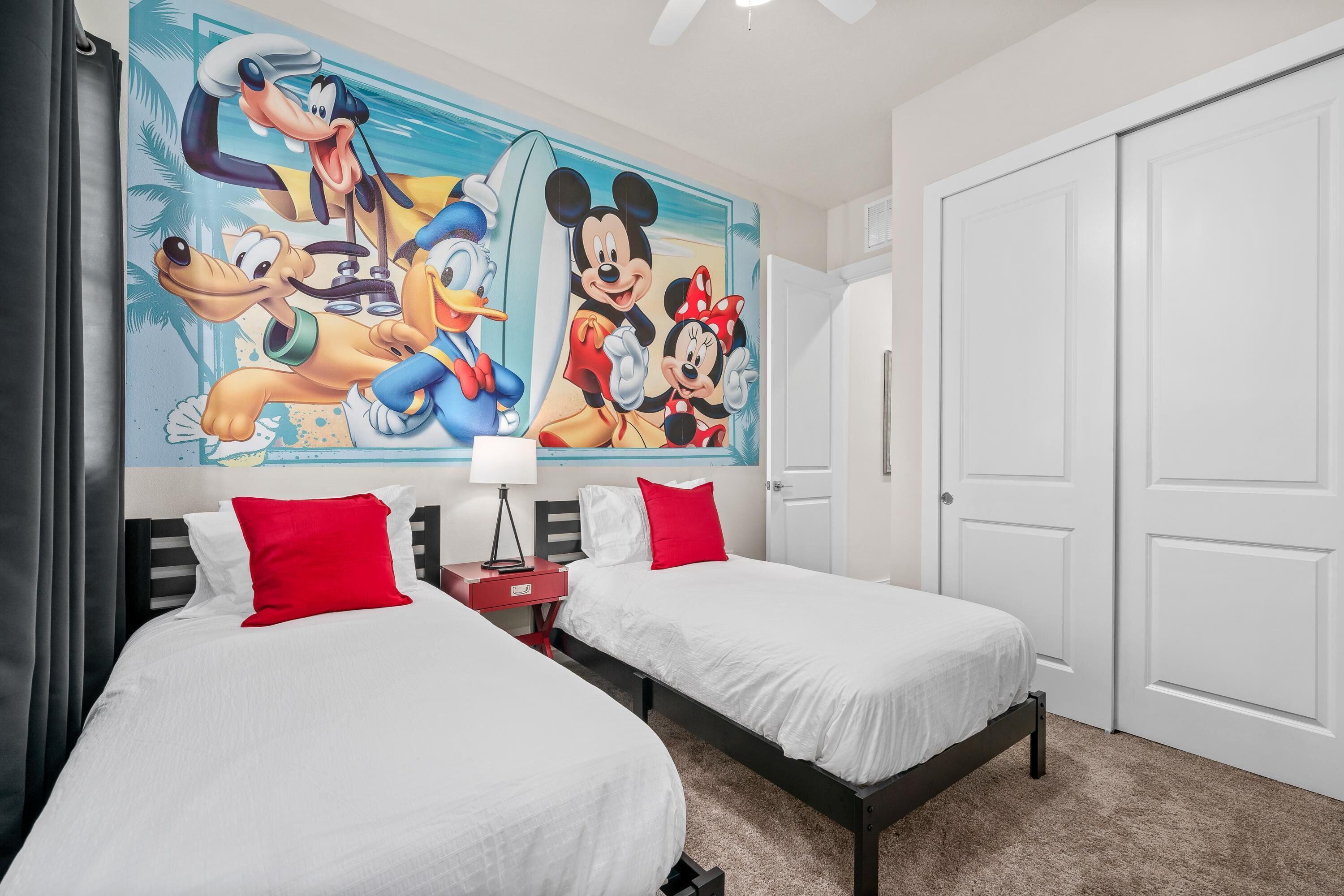 Quarto de hotel com duas camas, um travesseiro vermelho e branco, uma mesinha de cabeceira e um mural na parede com personagens de desenho animado.