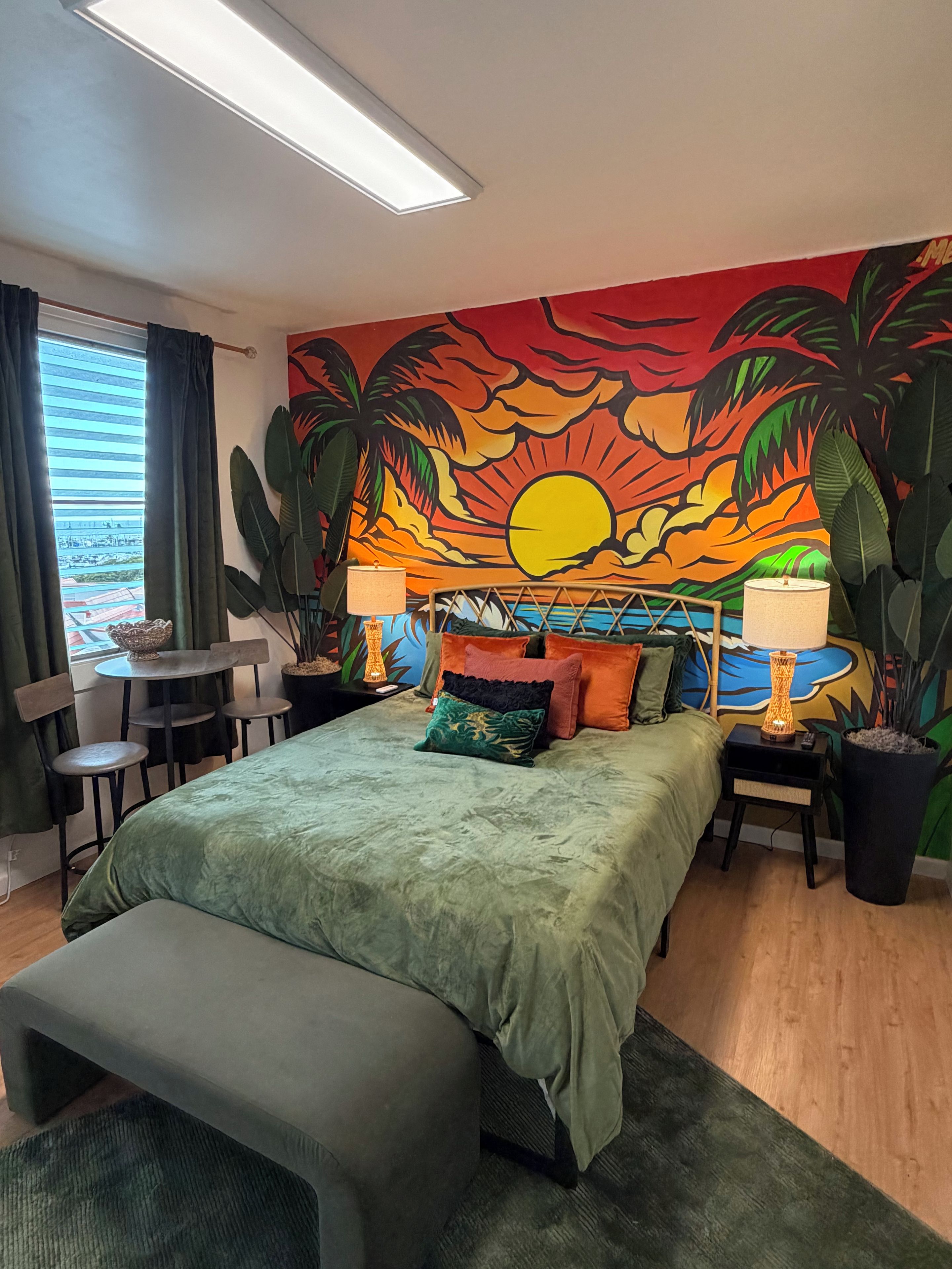 Quarto com cama, escrivaninha, cadeiras, janela com cortinas e um mural vibrante com pôr do sol na parede.