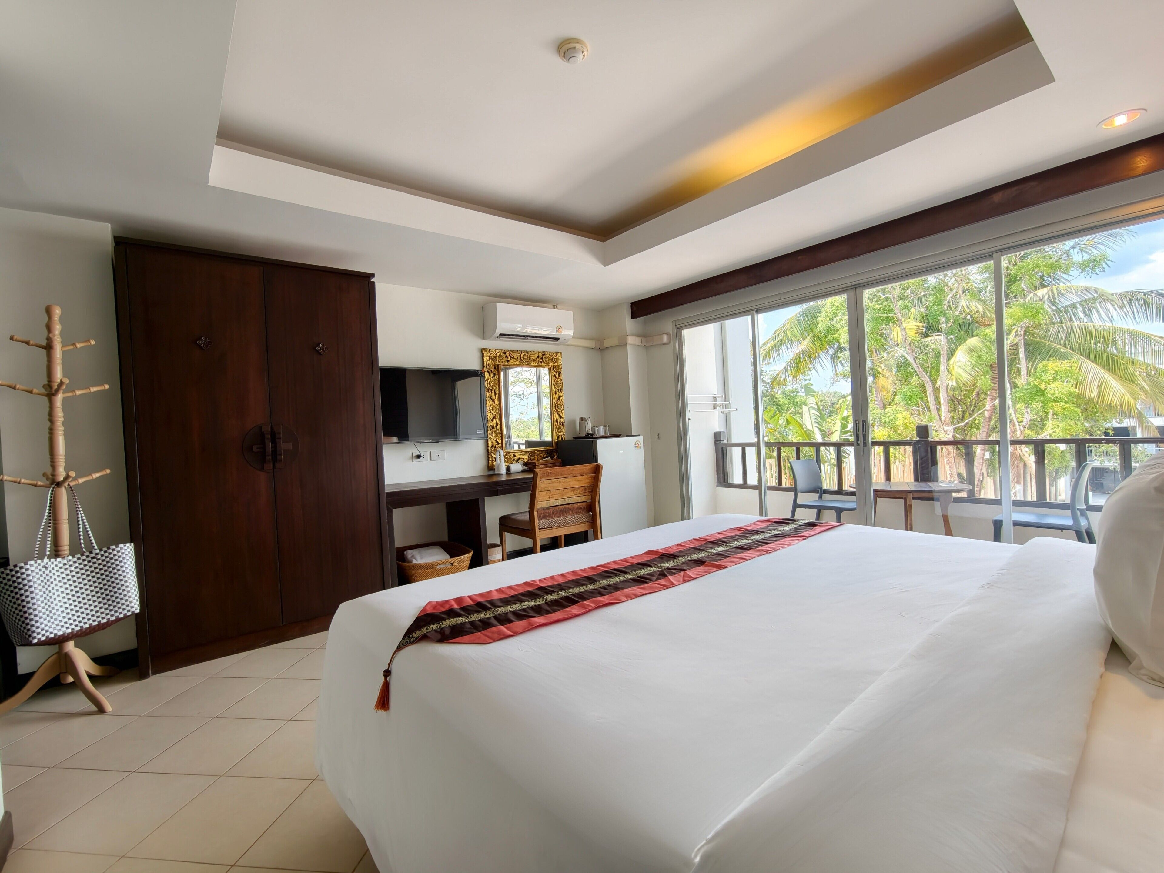 Quarto de hotel com cama, escrivaninha, cadeira, guarda-roupa, televisão e varanda com vista para palmeiras.