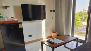 Quarto de hotel com televisão de tela plana na parede, um pequeno frigorífico, uma mesa de madeira com um comando remoto e um papel, e vista para o exterior através de uma janela grande.