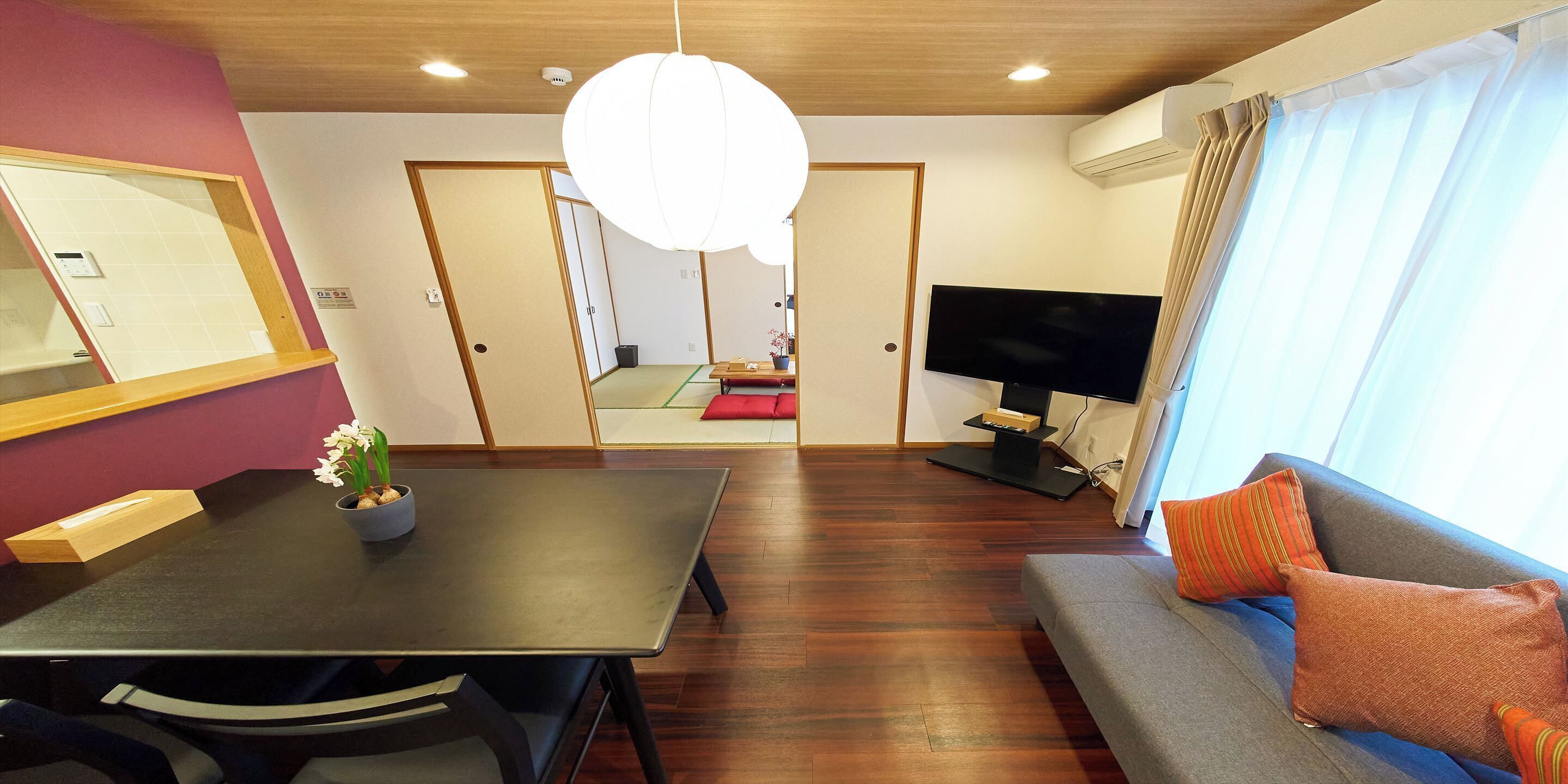 Uma sala de estar moderna com uma mesa de jantar, sofá, TV e vista para um espaço tradicional japonês ao fundo.