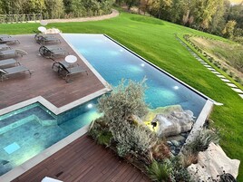 Uma piscina infinita moderna com deck de madeira e espreguiçadeiras, cercada por gramado bem cuidado e um caminho de pedra.