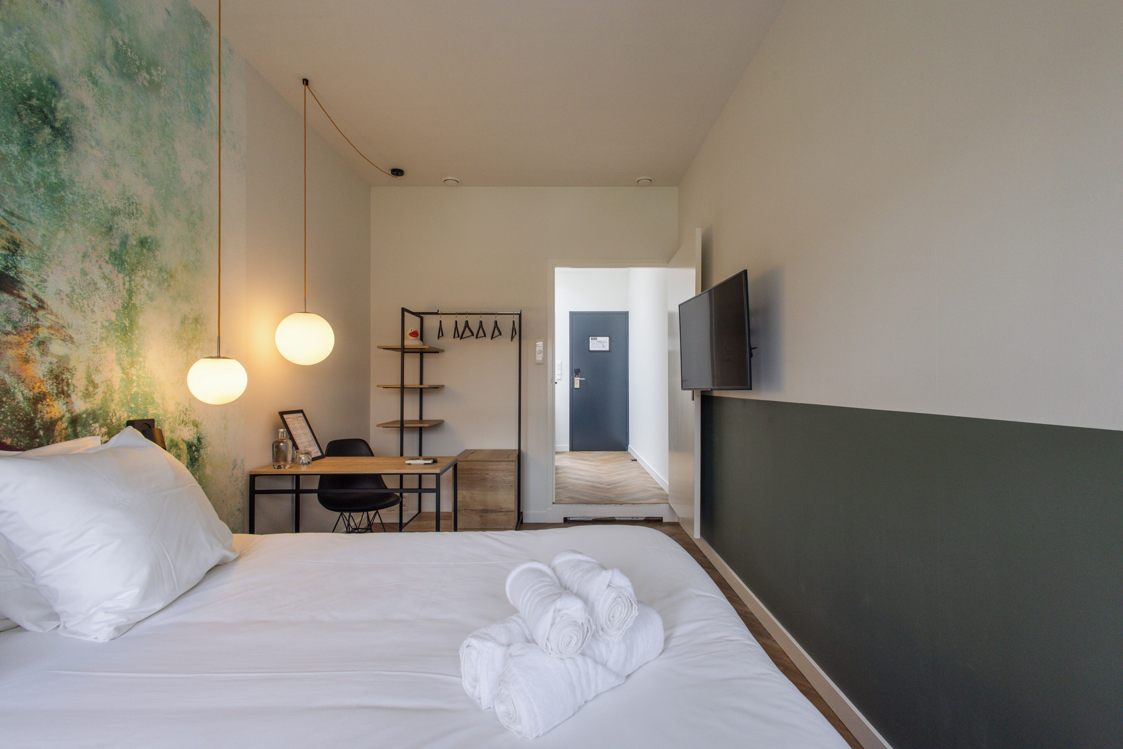 Quarto de hotel com cama, escrivaninha, cadeira, televisão e um quadro na parede.