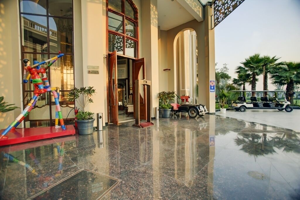 Entrada de hotel com uma escultura vermelha e branca, porta de vidro e área de estacionamento com palmeiras.