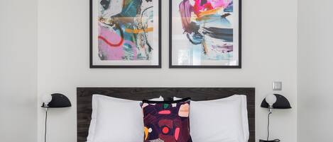 Quarto de hotel com cama, duas pinturas abstratas emolduradas, luminária moderna e almofada decorativa.