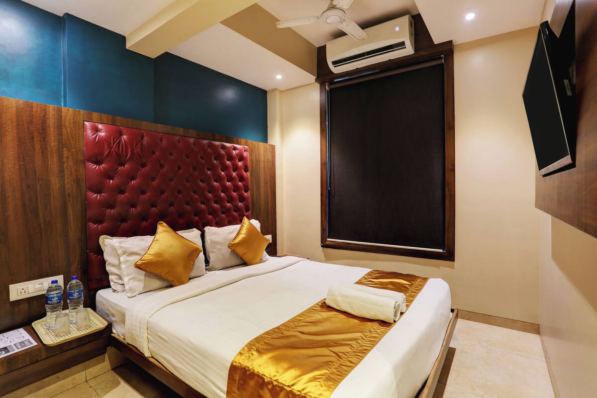 Quarto de hotel com cabeceira estofada vermelha, uma janela grande e uma TV de tela plana fixada na parede.