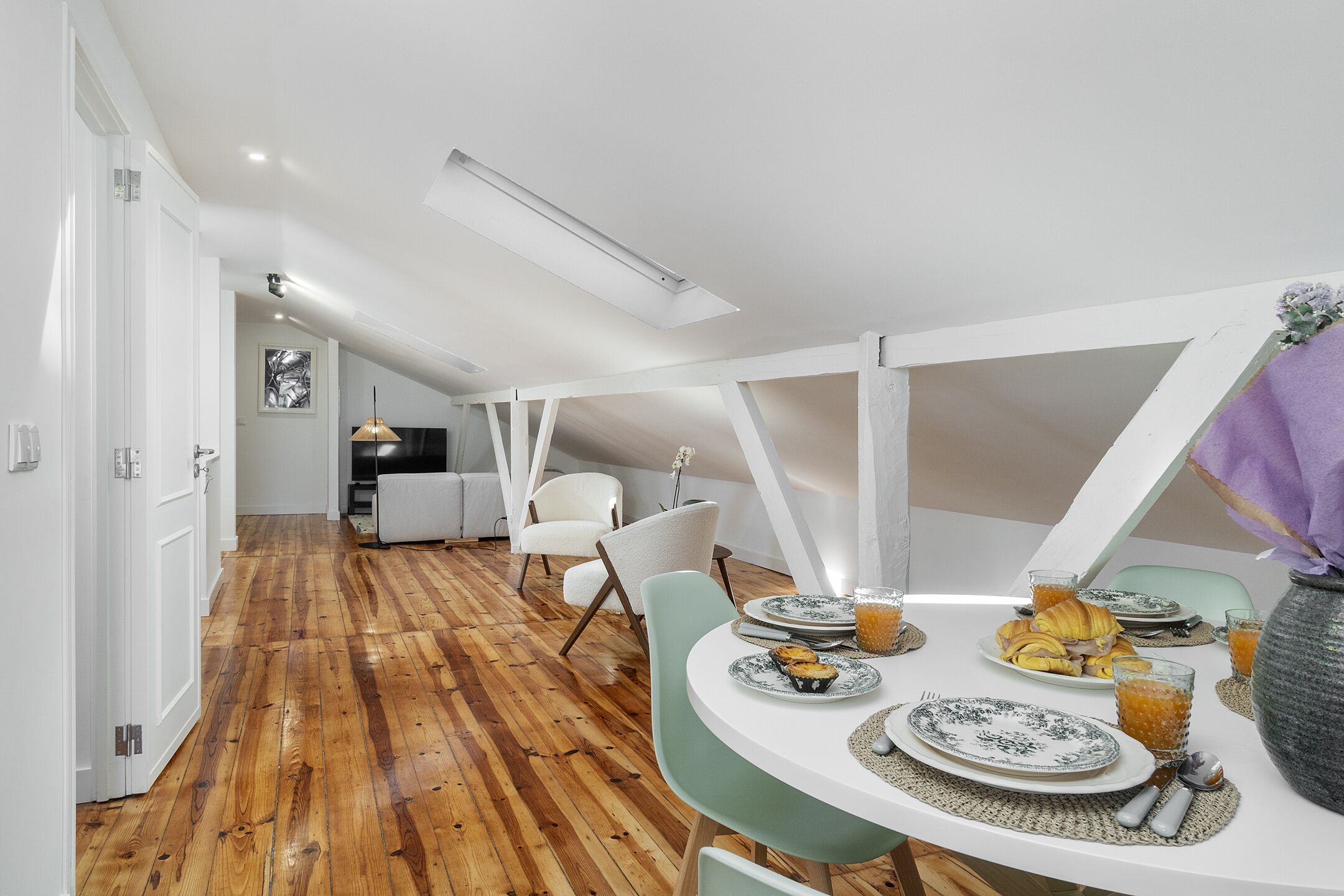 Uma sala de jantar com chão de madeira, paredes brancas e uma mesa posta com pratos, copos e um vaso.