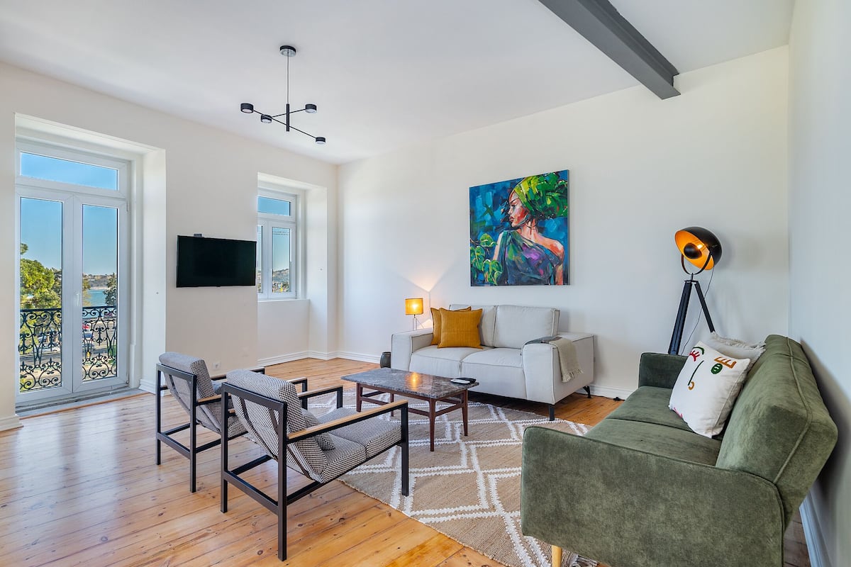 Uma sala de estar moderna com um sofá, uma mesa de centro e uma televisão. Há um quadro na parede e um abajur na mesinha lateral.