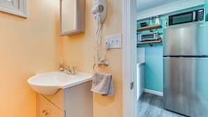 Casa de banho com lavatório, porta-toalhas, secador de cabelo, espelho e uma porta que dá para uma cozinha com frigorífico e micro-ondas.