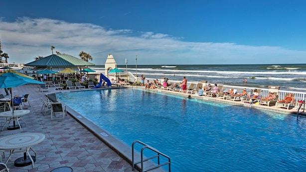 Uma área de piscina com espreguiçadeiras e guarda-sóis, um pequeno edifício com telhado verde e vista para o oceano.