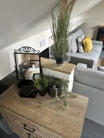 Baú de madeira com plantas, um espelho e um sofá cinzento com almofada amarela numa sala de estar.
