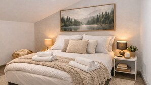 Um quarto com uma cama grande, duas mesas de cabeceira, um quadro na parede e uma pequena prateleira com livros e uma vela.