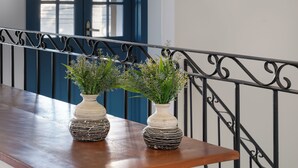Uma mesa de madeira com dois vasos brancos contendo plantas, uma porta azul e um guarda-corpo de ferro forjado preto.