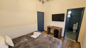 Quarto com cama, mesa de cabeceira, televisão e banheiro visível por uma porta aberta.