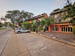Vista da rua com uma caminhonete branca estacionada na lateral, um edifício com a placa 'Samui Hi Samui Hotel & Restaurant' e uma calçada arborizada.