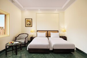 Quarto de hotel com duas camas, uma cadeira, uma mesinha com frutas e um quadro na parede.