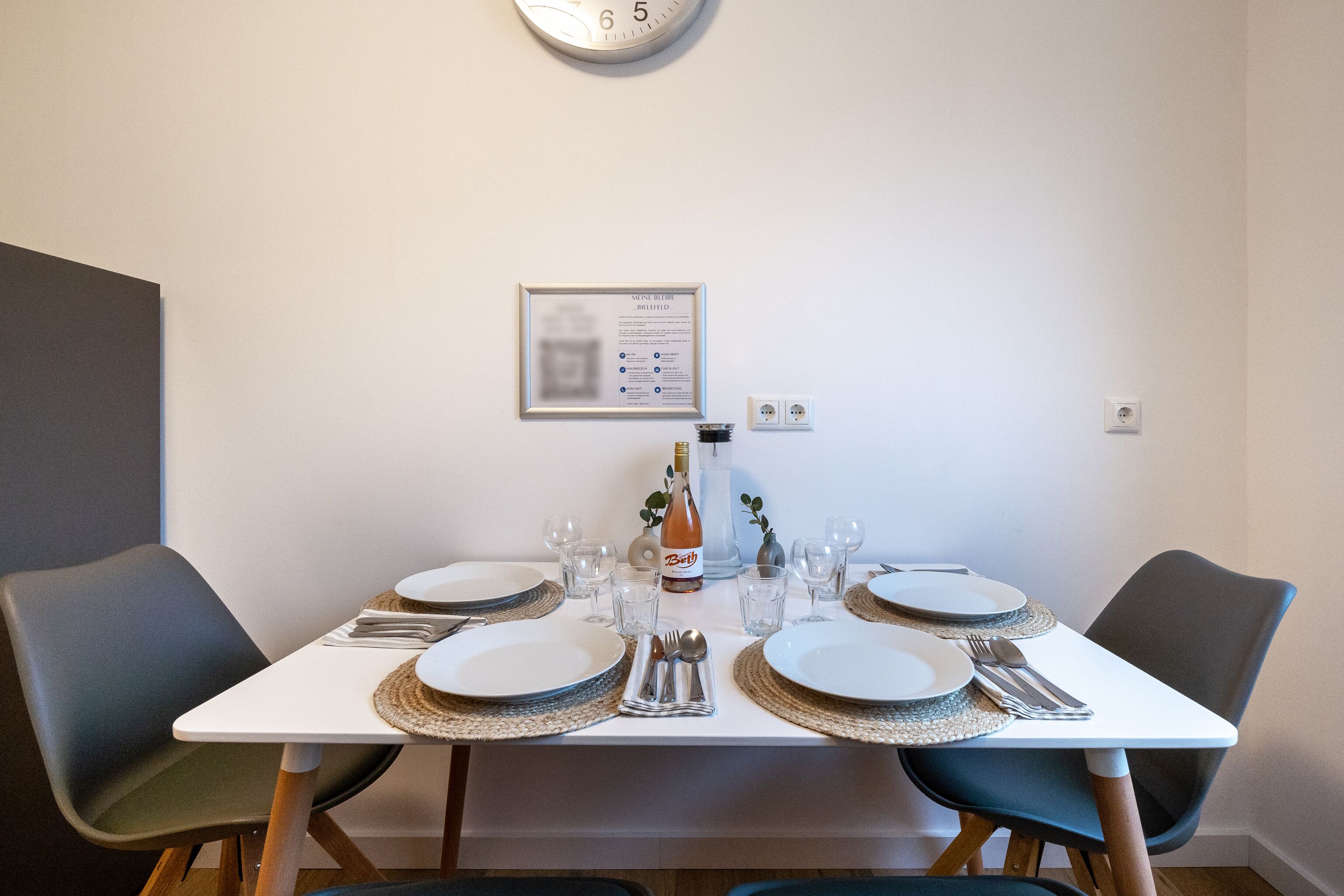 Uma mesa de jantar posta com pratos, talheres e copos, com uma garrafa de vinho e um aviso emoldurado na parede.