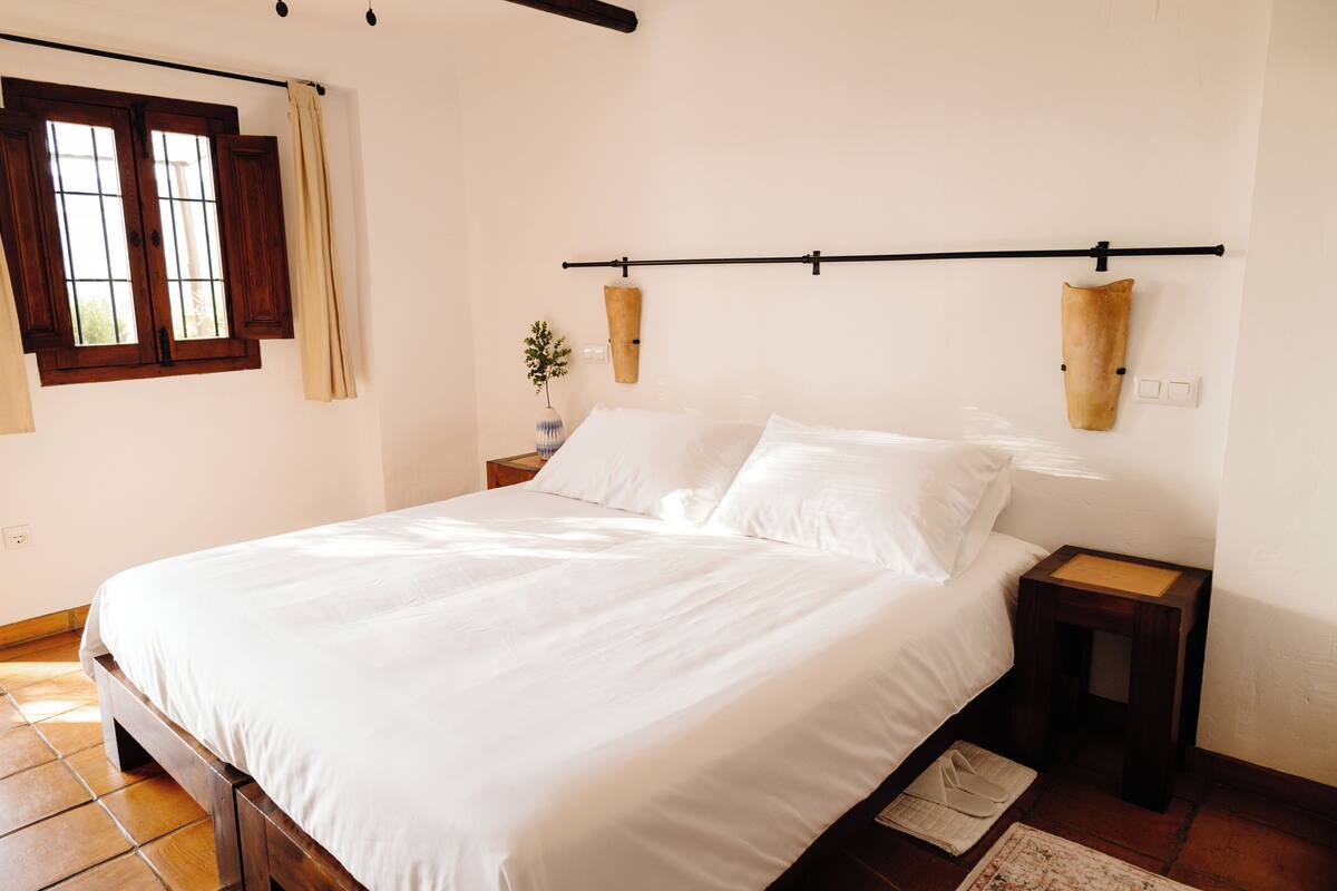 Uma cama bem arrumada com roupa de cama branca, cabeceira de madeira, mesa de cabeceira com abajur e janela com cortinas.