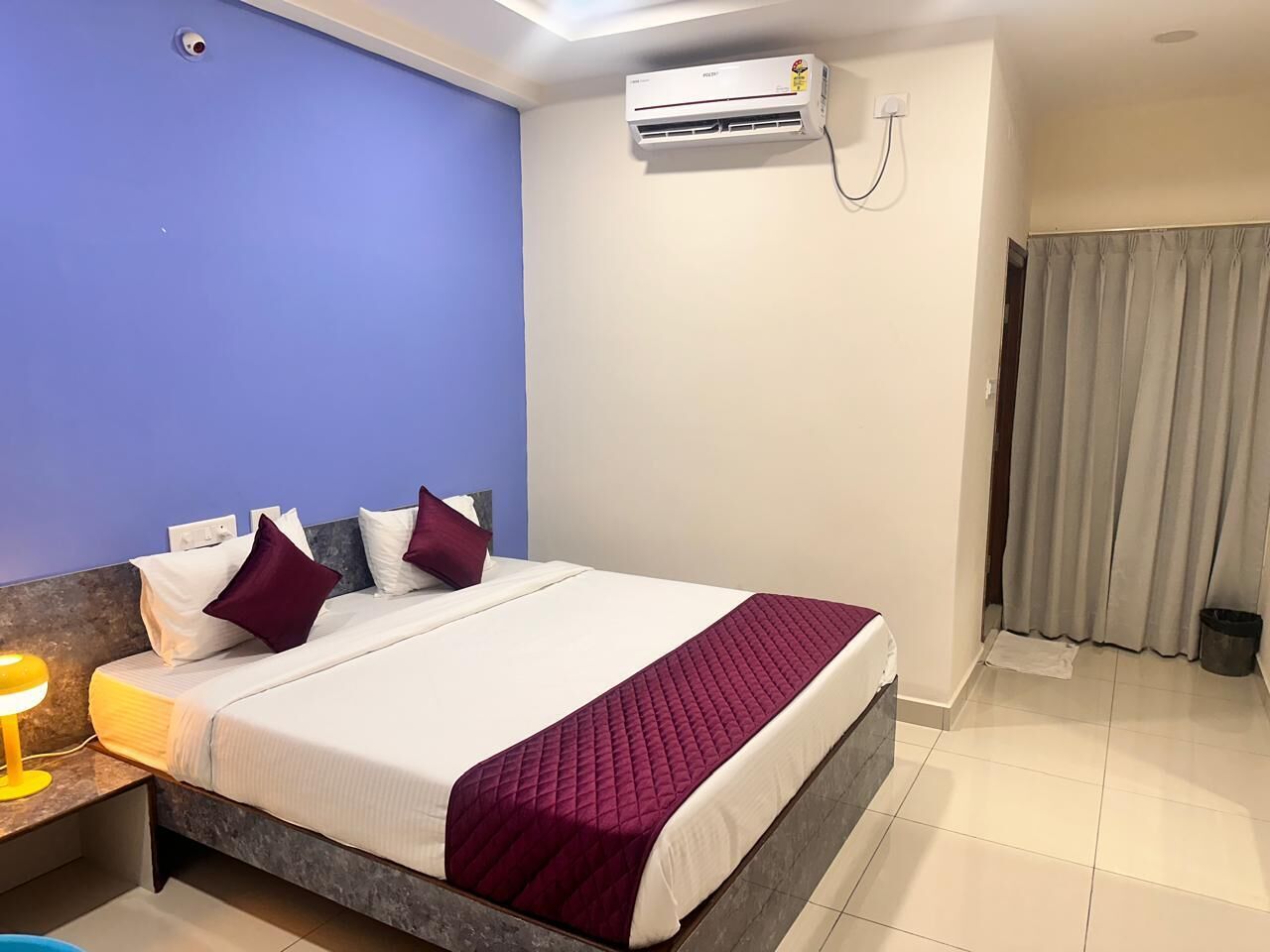 Cama bem arrumada com colcha branca e avelã, uma luminária de cabeceira e ar condicionado fixado na parede.