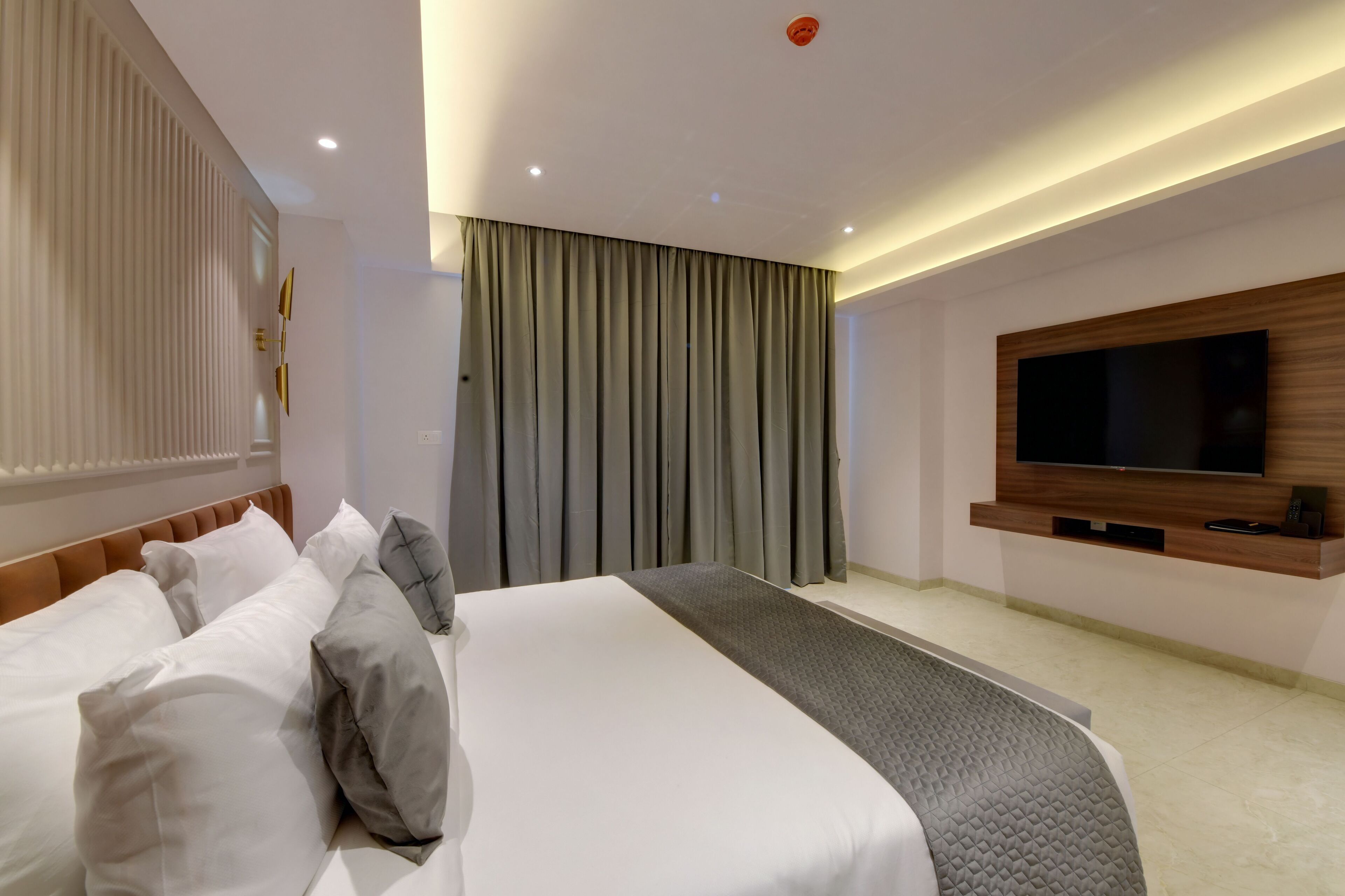 Quarto de hotel moderno com uma cama grande, televisão de tela plana na parede e uma janela ampla com cortinas.