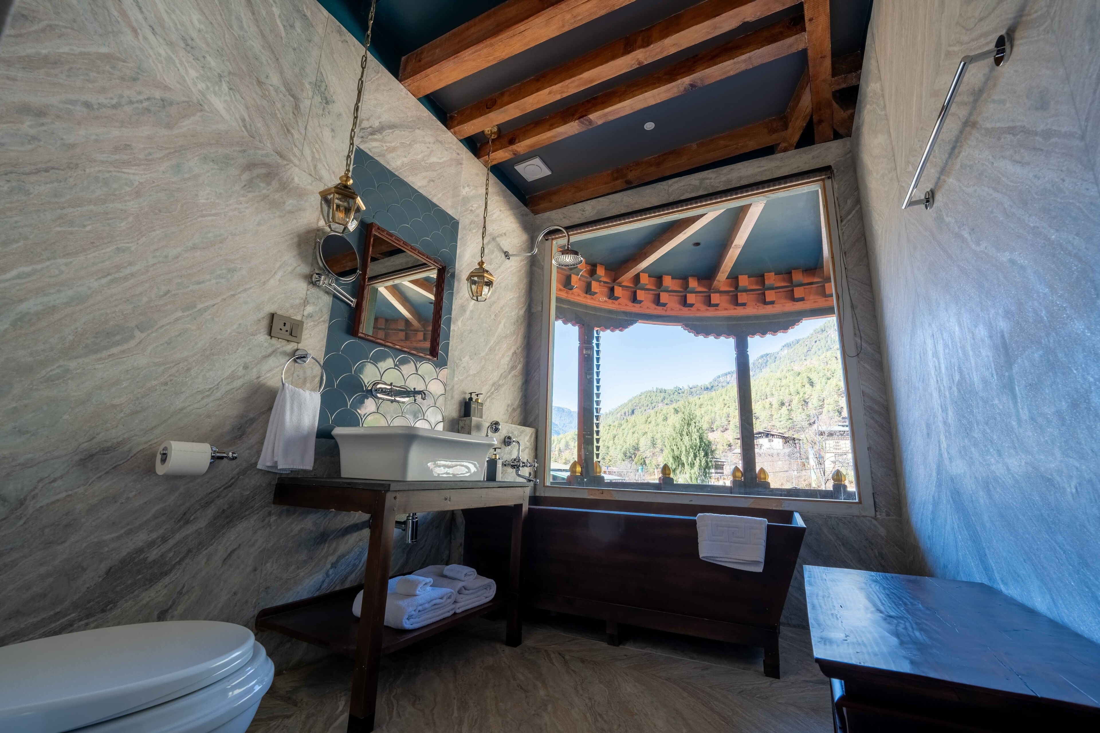 Casa de banho com parede de mármore, vigas de madeira, lavatório, sanita e janela com vista para o exterior.