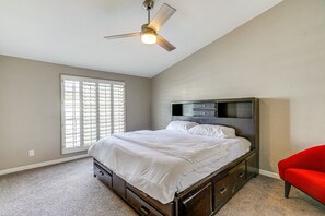 Quarto com cama, cadeira vermelha, ventoinha no teto e janela com persianas.