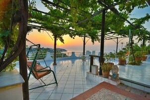 Terraço no telhado com cadeira de balanço, plantas em vasos e vista do pôr do sol sobre uma ponte.