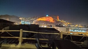 Vista noturna de uma cidade com uma colina imponente iluminada em laranja, uma mesquita com um minarete e uma ponte com guarda-corpo.