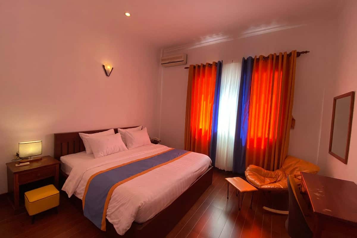 Quarto de hotel com cama, mesa de cabeceira, luminária, banco e uma cadeira.