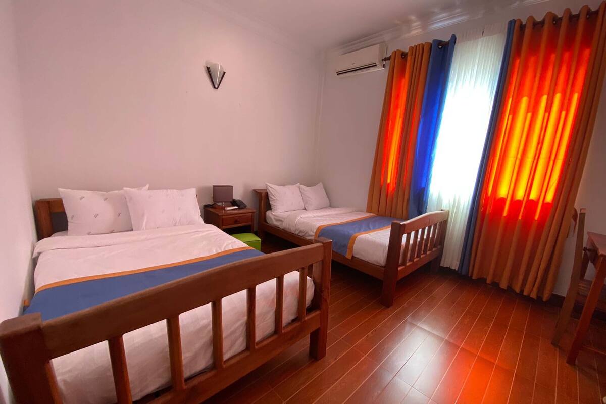 Quarto de hotel com duas camas de madeira, uma mesinha de cabeceira, uma caixa de armazenamento verde e uma janela com cortinas coloridas.