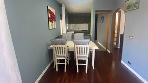 Uma área de refeições com uma mesa e cadeiras brancas, uma luminária no teto e uma cozinha ao fundo.