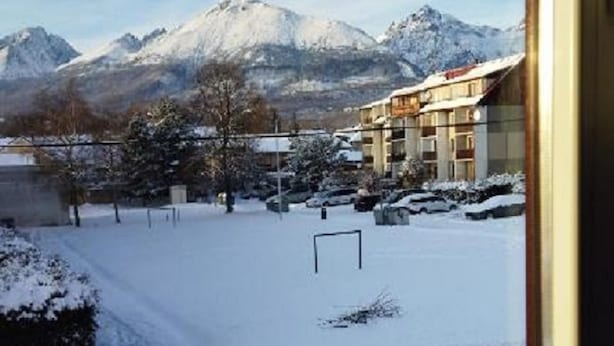 Paisagem nevada com montanhas ao fundo, céu limpo e um edifício com telhado coberto de neve.
